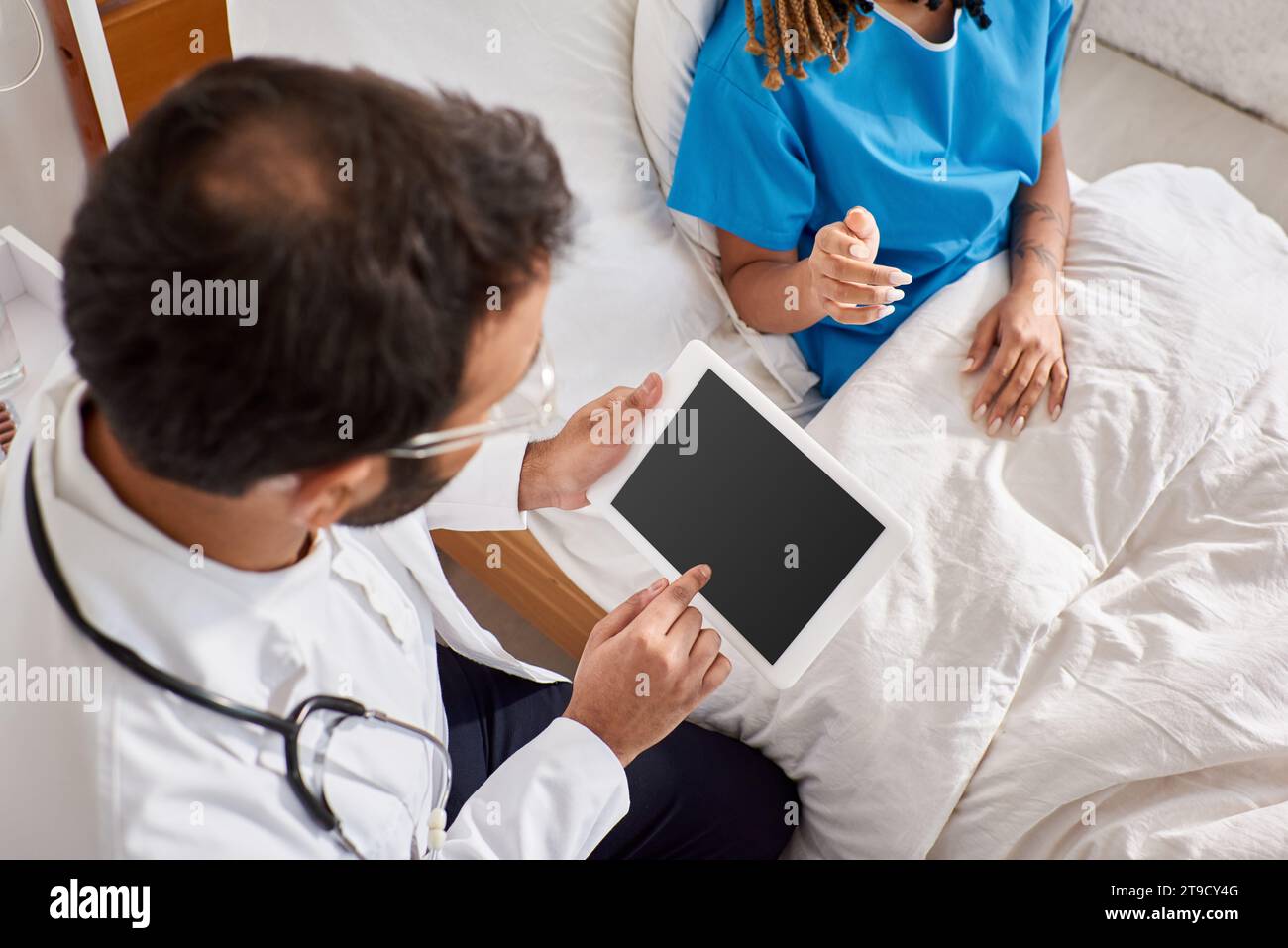 african american patient sitting on bed near her indian doctor taking ...