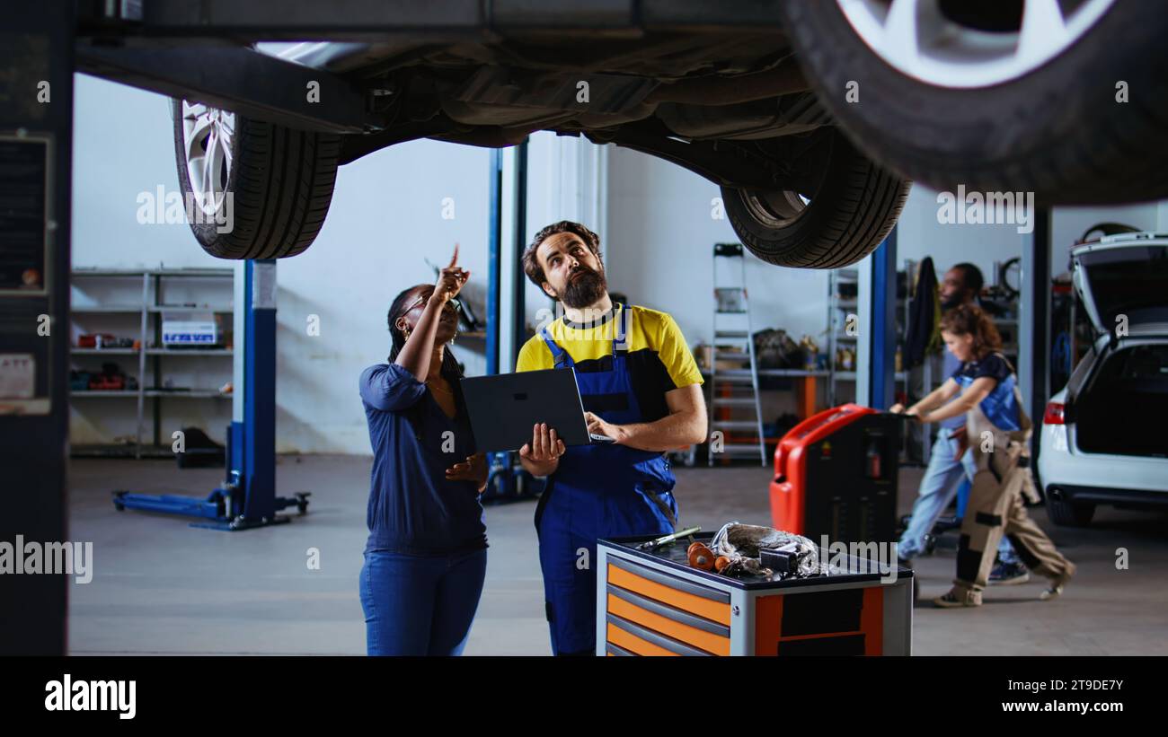 Serviceman and customer standing underneath car in garage using laptop ...