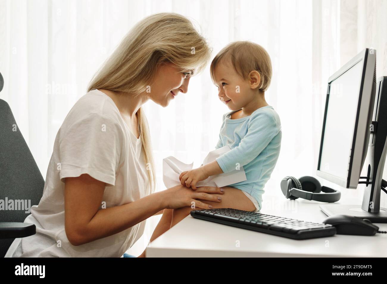 Playful little boy sitting on the desk at his freelancer mother's home ...