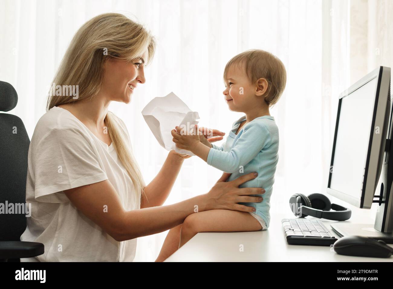 Playful little boy sitting on the desk at his freelancer mother's home ...