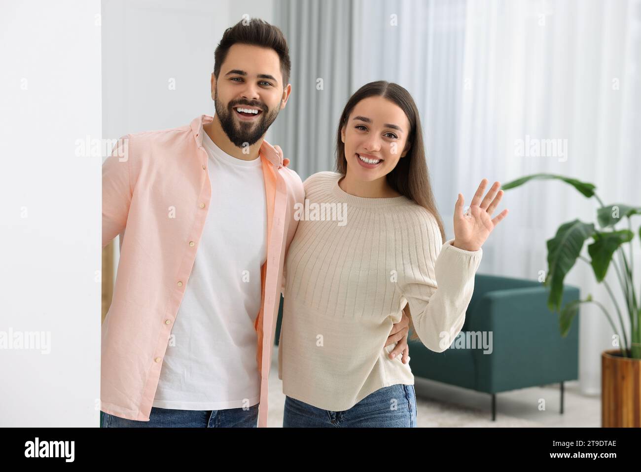 Happy couple waving near white wall at home. Invitation to come in room ...