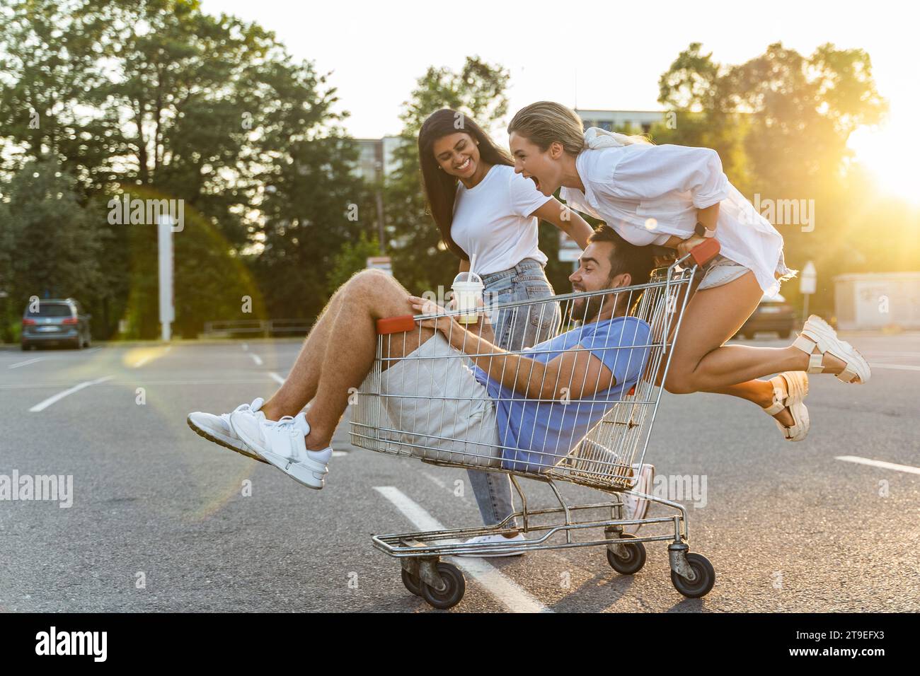 Three diverse friends having fun and riding shopping cart on parking ...