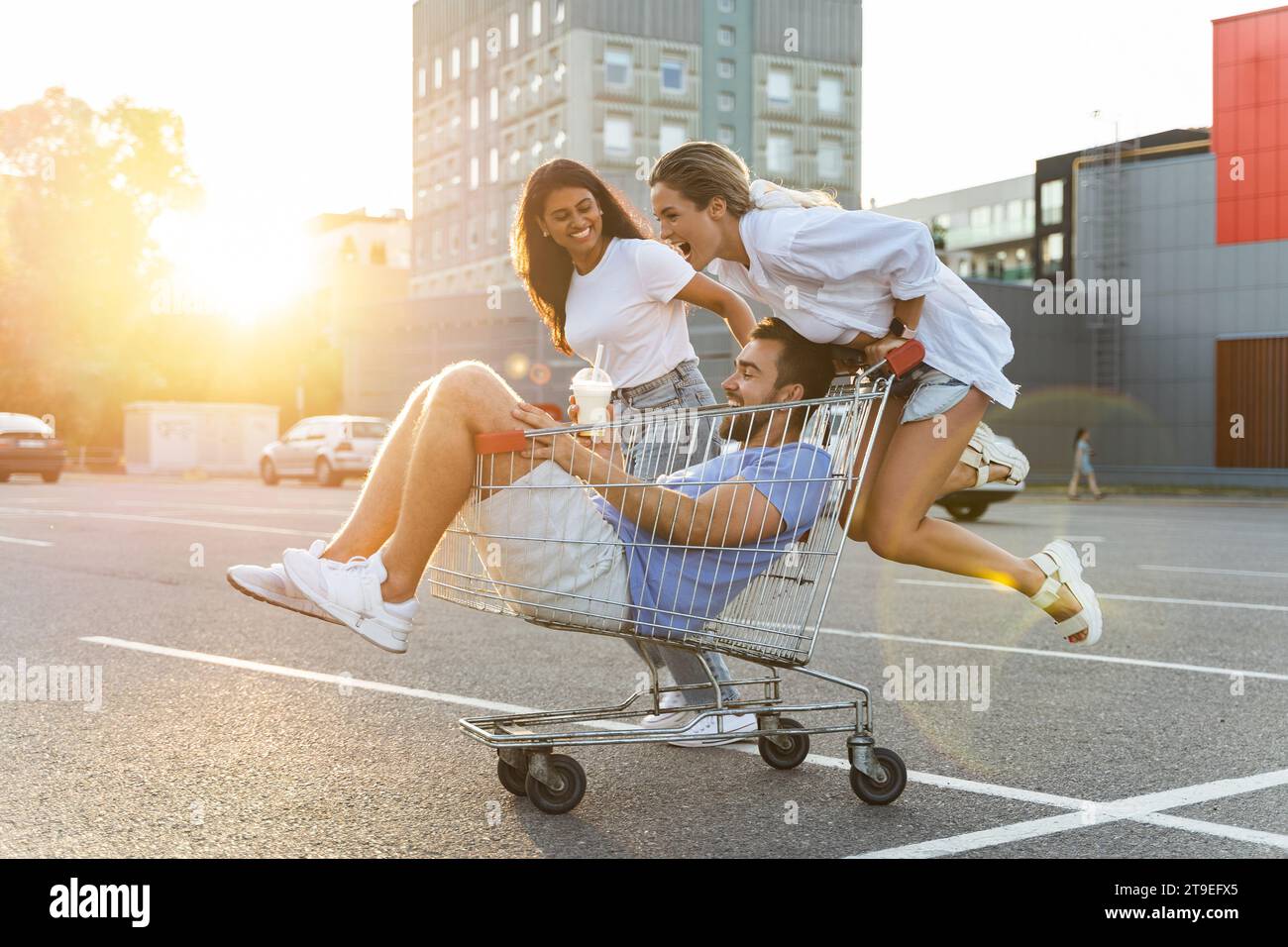 Three diverse friends having fun and riding shopping cart on parking ...