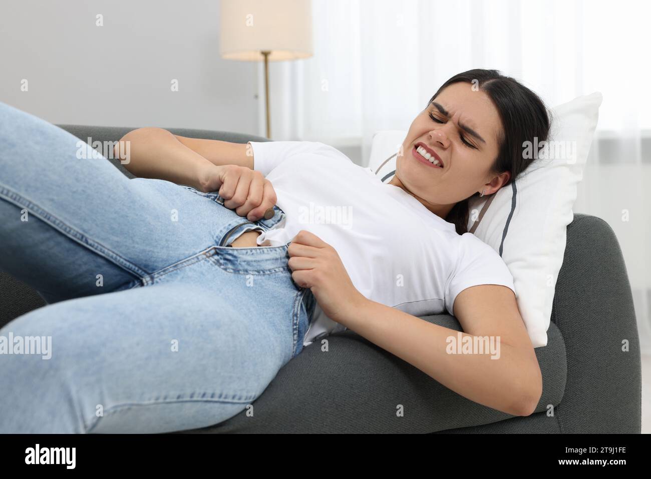 Young Woman Struggling To Squeeze Into Tight Jeans While Lying On Sofa
