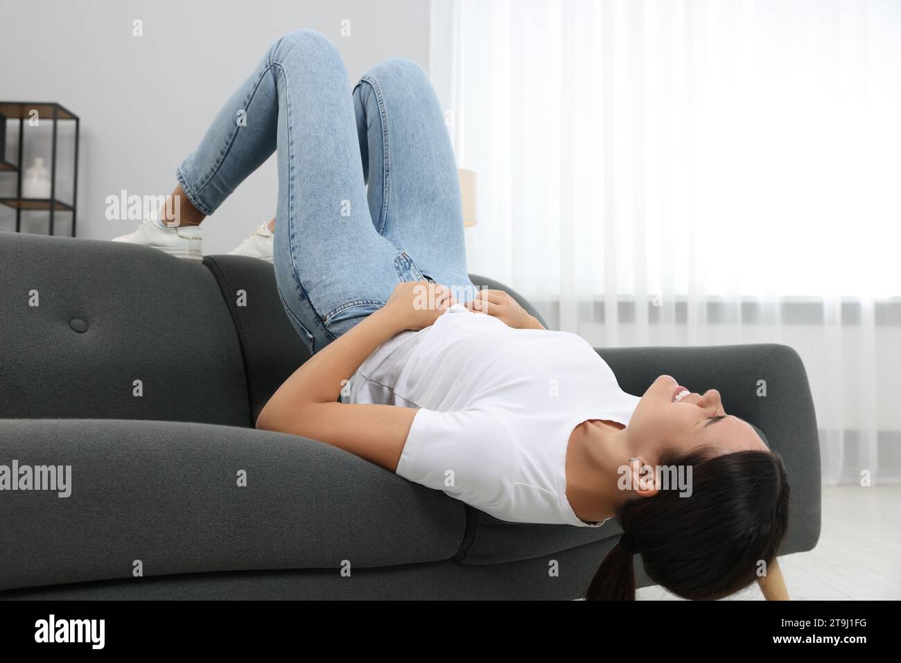 Young woman struggling to squeeze into tight jeans while lying on sofa ...