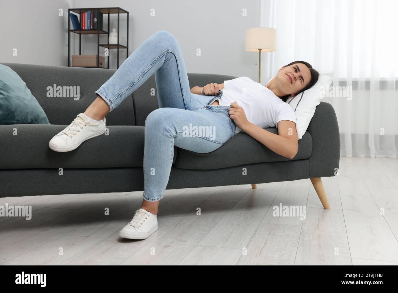 Young Woman Struggling To Squeeze Into Tight Jeans While Lying On Sofa