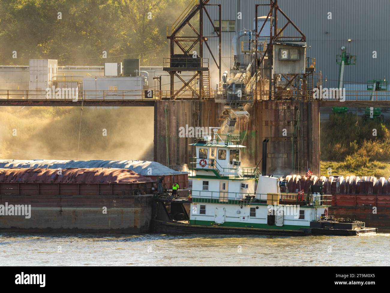 Tugboat pushing freight barges past grain loading dock with dust ...