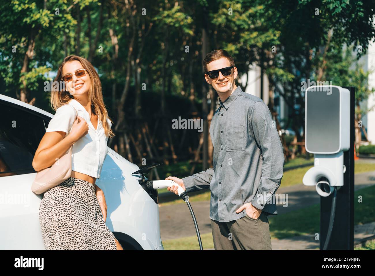Young couple travel with EV electric car charging in green sustainable city outdoor garden in ...
