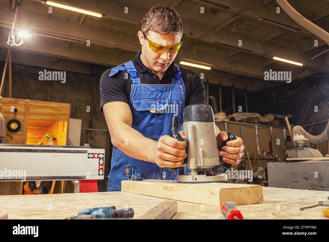 A young man wearing safety goggles carpenter builder pre-works the edges of the wooden board ...