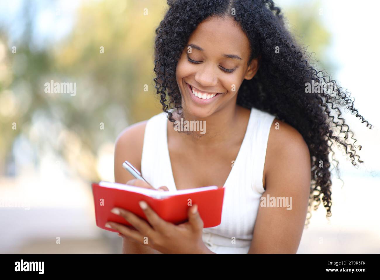 Front view portrait of a happy black woman writing on agenda outdoors ...