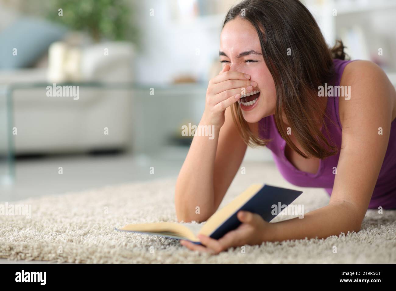 Woman laughing loud reading a paper book lying on the floor at home ...