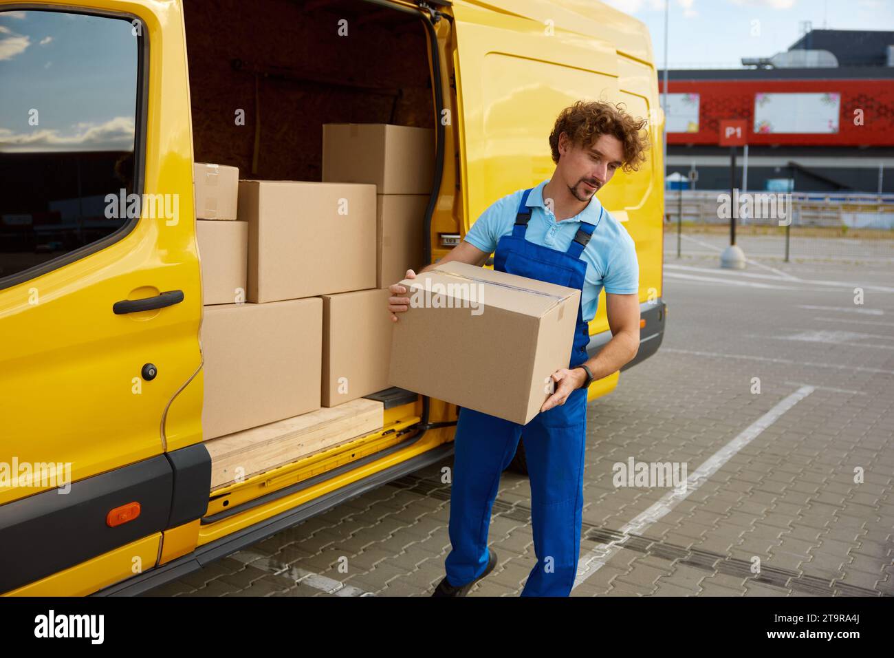Young deliveryman carrying cardboard box unloading minivan on street ...