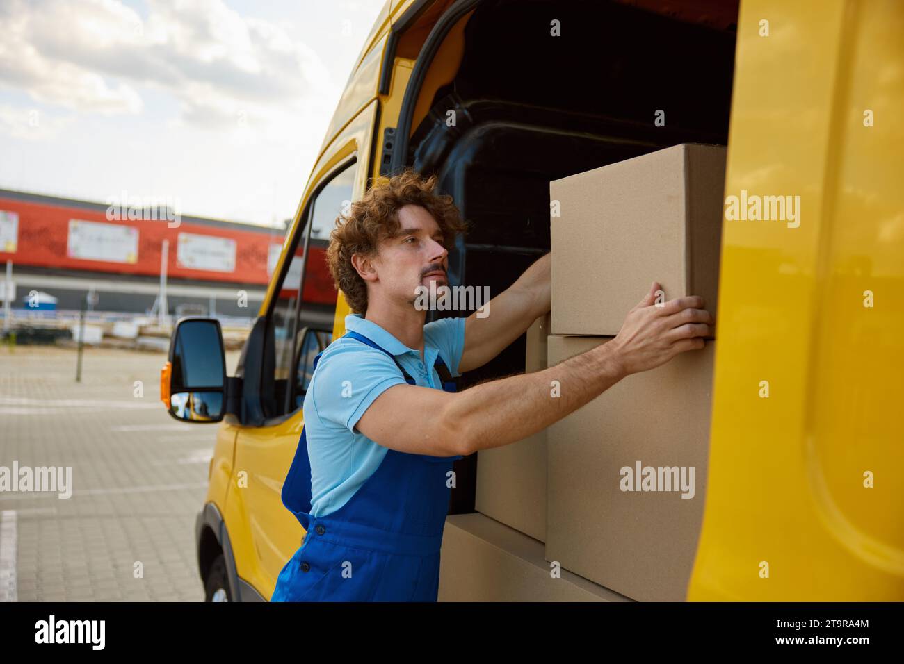 Young deliveryman carrying cardboard box unloading minivan on street ...
