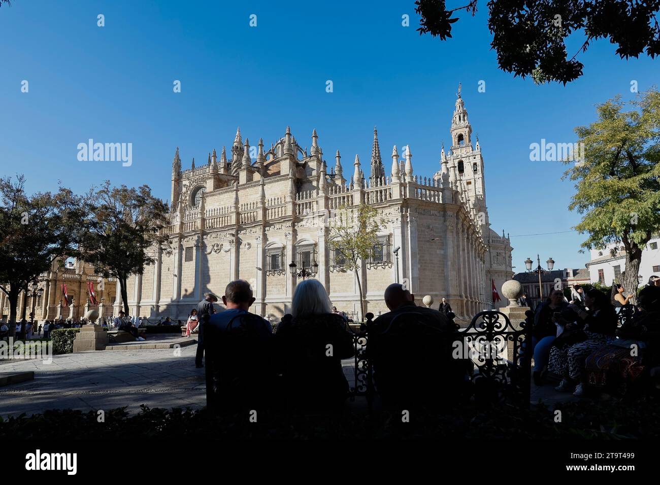 People sitting on a bench in the shade looking at the Seville Cathedral ...