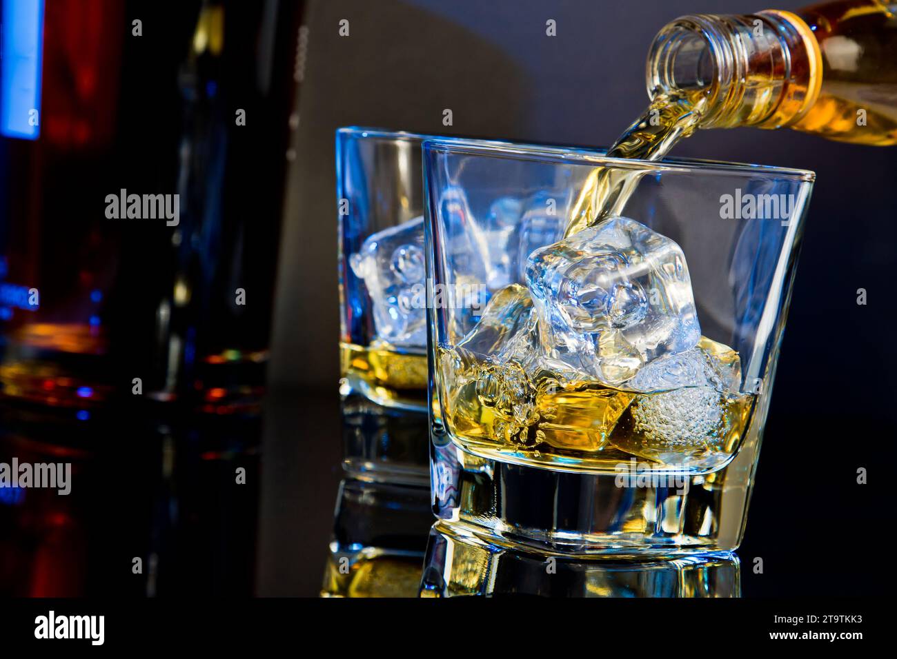 barman pouring whiskey in two glasses with ice cubes on table with light tint blue and ...