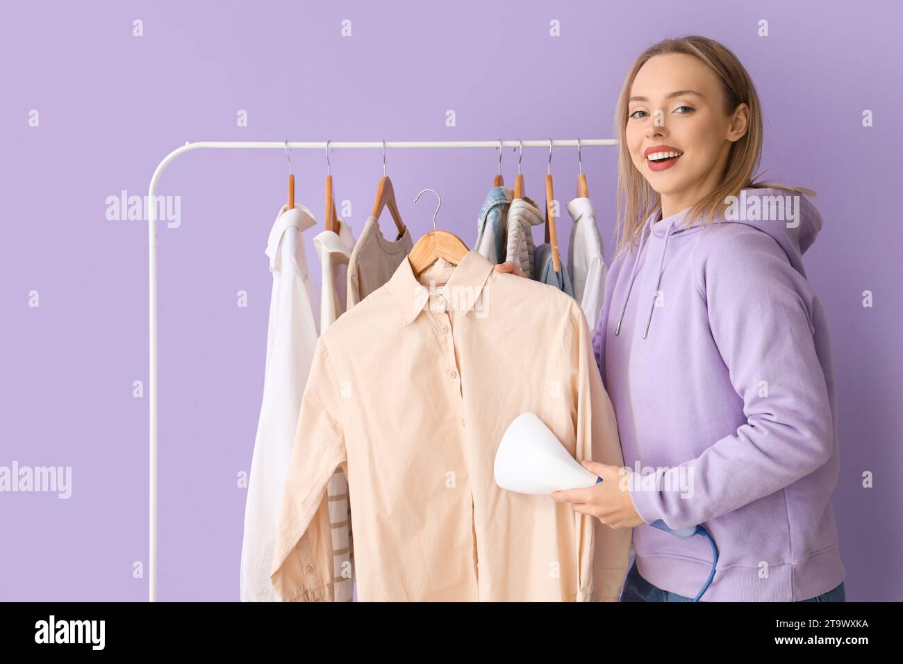 Pretty young woman steaming clothes with modern steamer on lilac ...