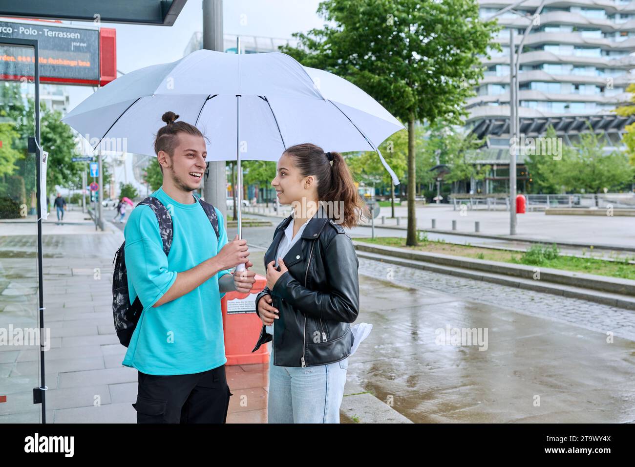 Teenagers guy and girl at bus stop under an umbrella waiting for city ...