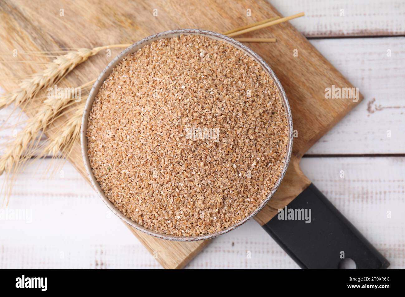 Dry wheat groats in bowl and spikelets on white wooden table, top view ...