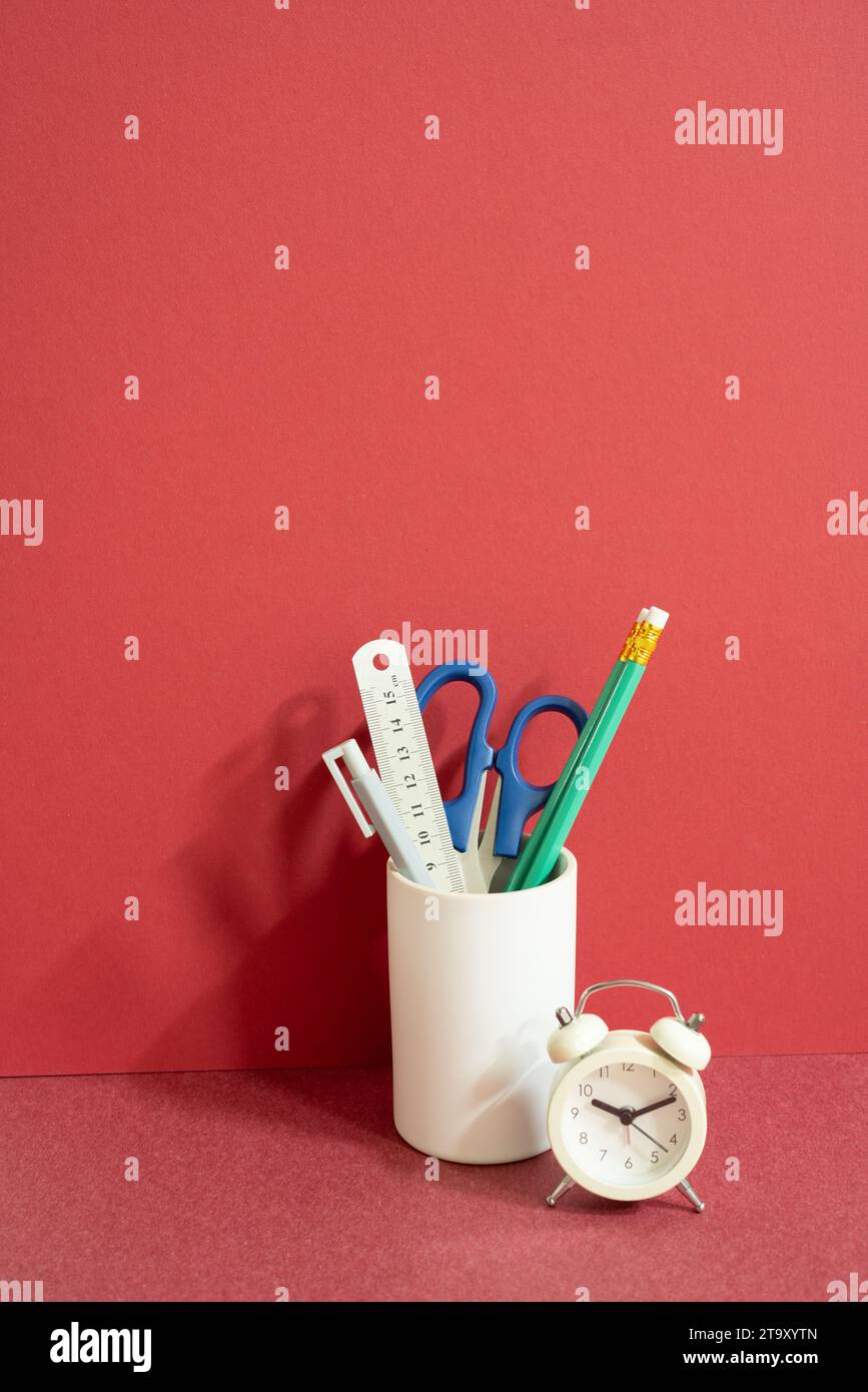 Pencil holder with ruler, scissors, pen and alarm clock on red desk ...