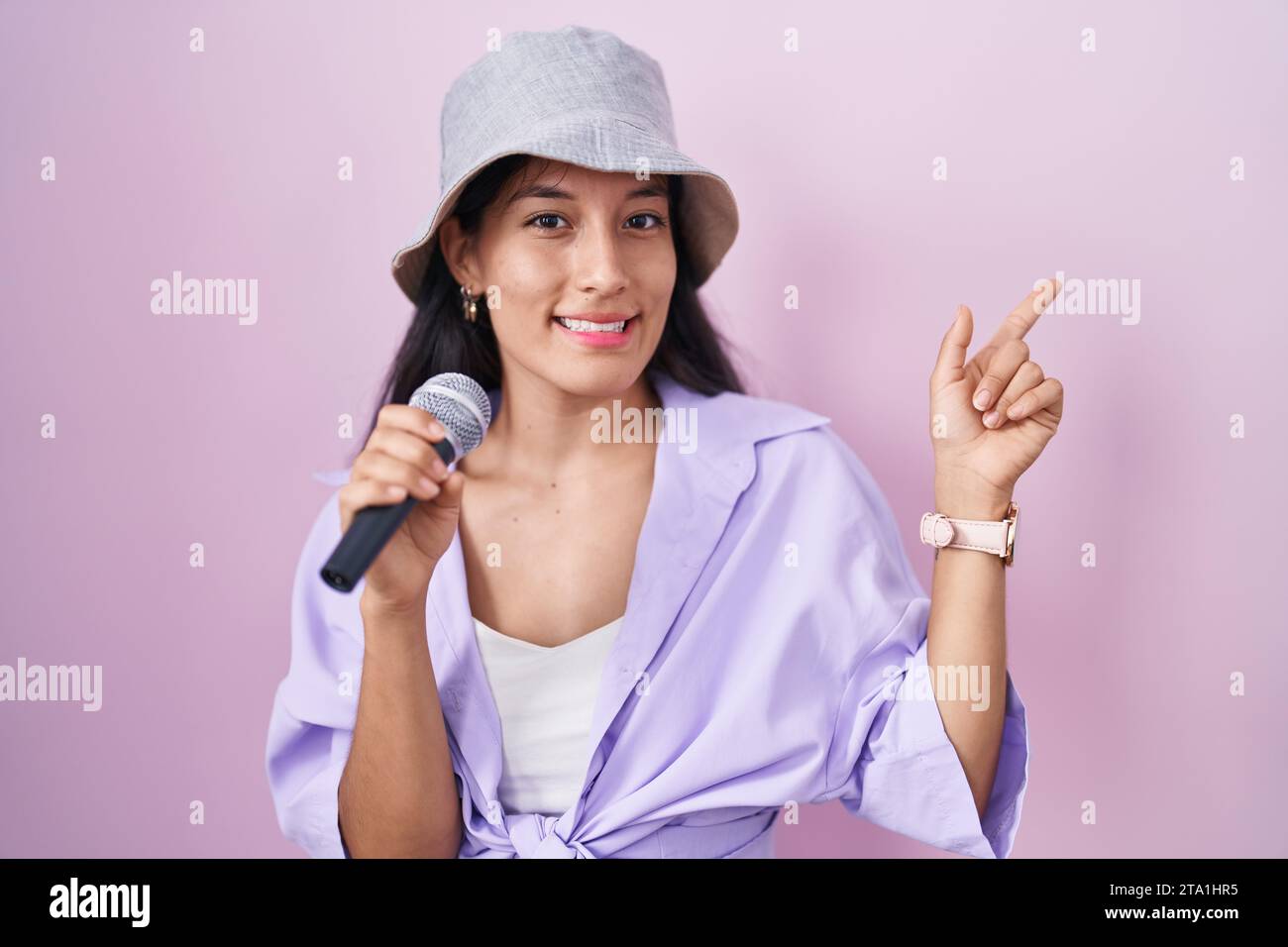 Young hispanic woman singing song using microphone over pink background ...