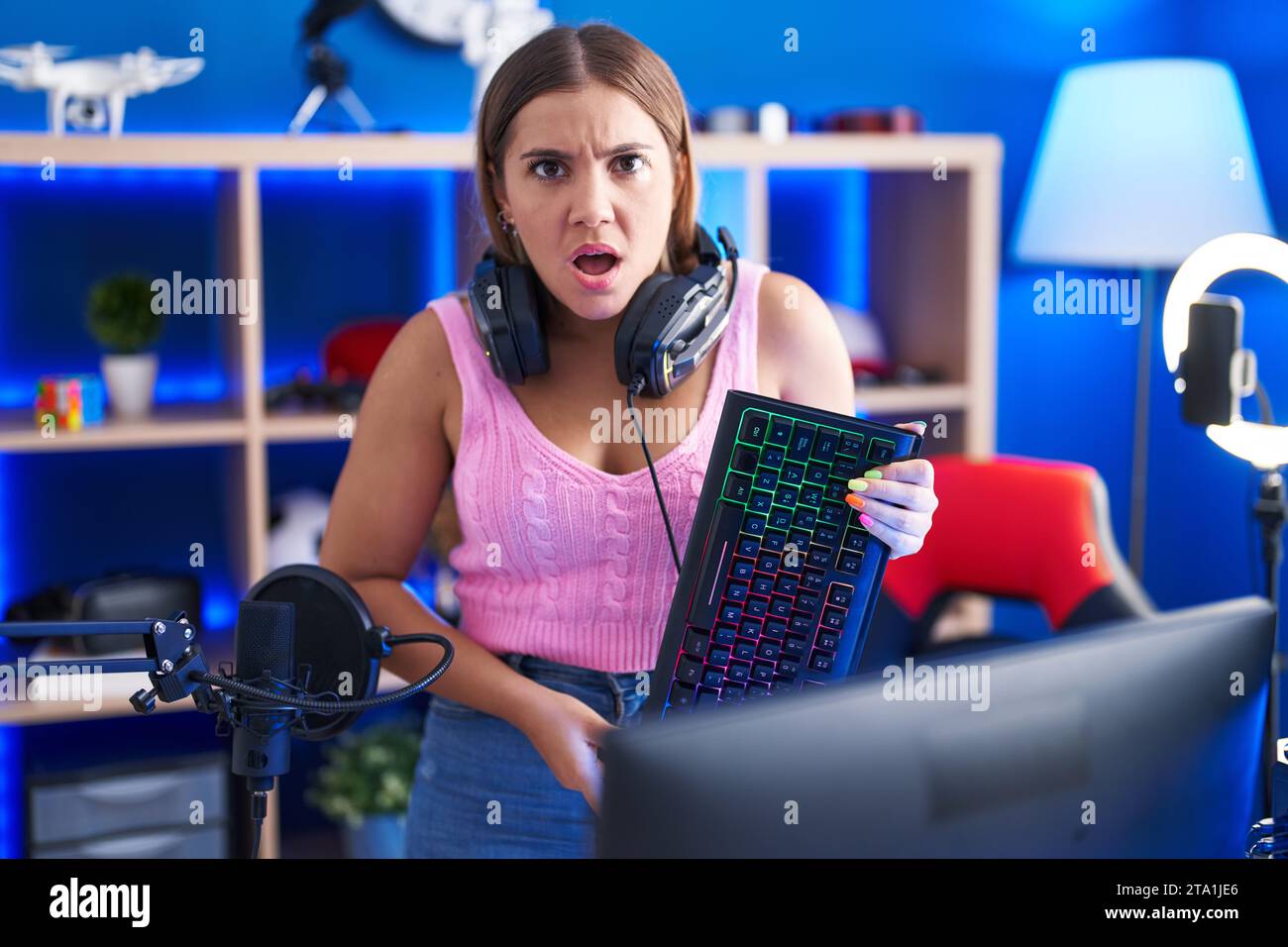 Young blonde woman playing video games holding keyboard in shock face ...