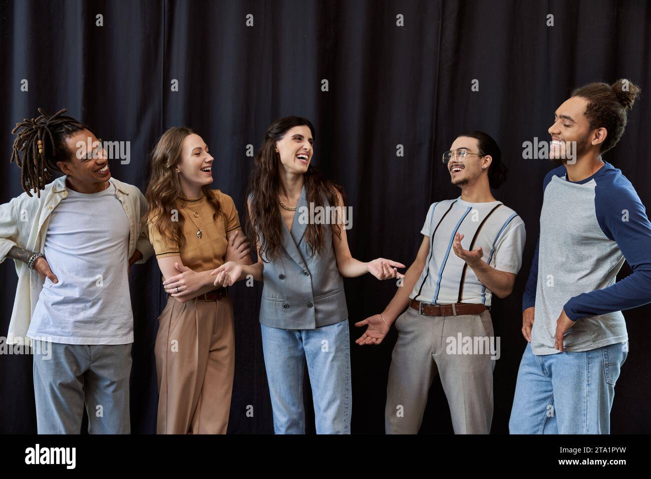 cheerful multicultural colleagues posing and laughing near black drape ...
