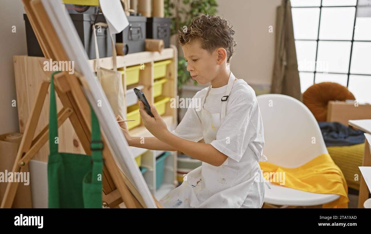 Adorable blond boy artist earnestly looking at smartphone, learning to ...
