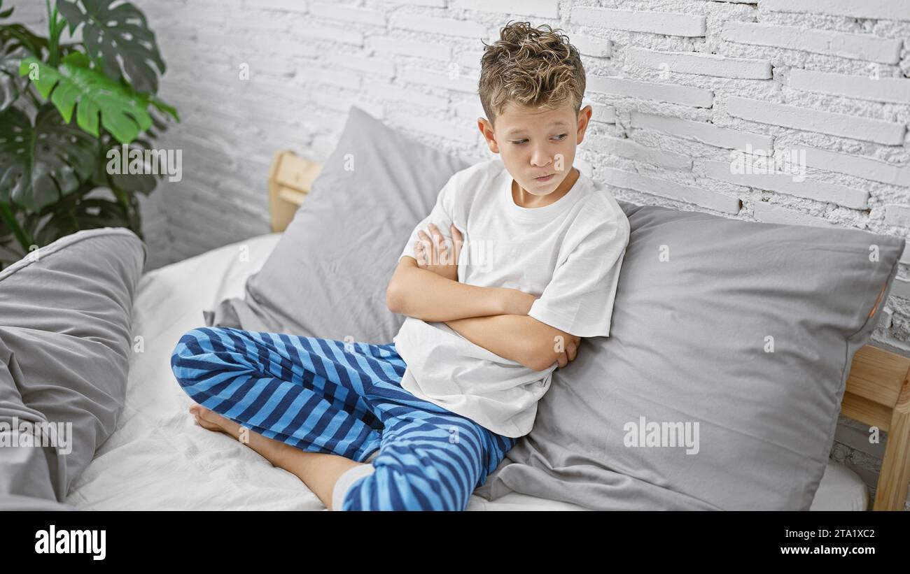 Adorable blond boy, sitting on bed with arms crossed, looking upset in ...