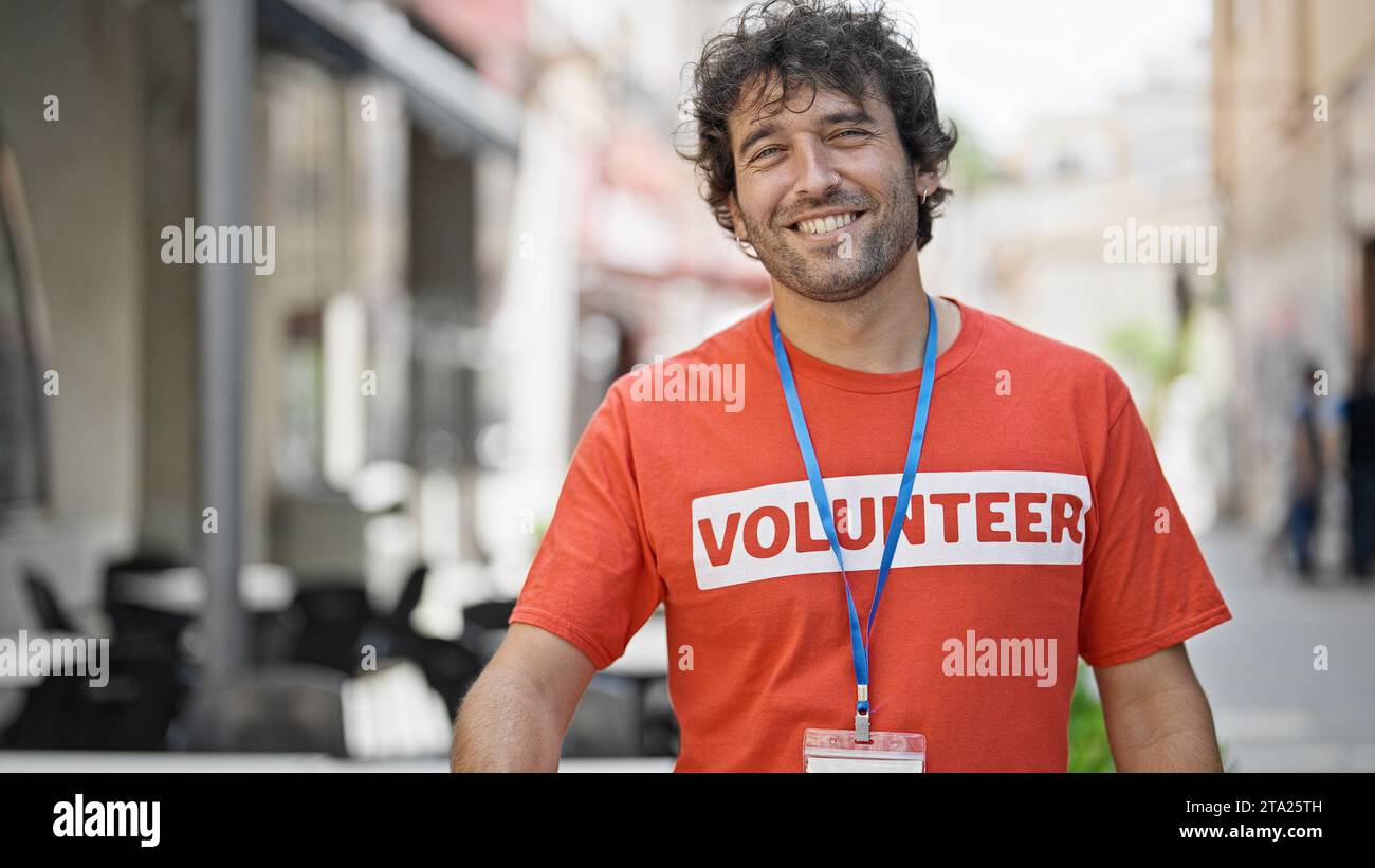 Young hispanic man activist wearing volunteer uniform smiling at street ...