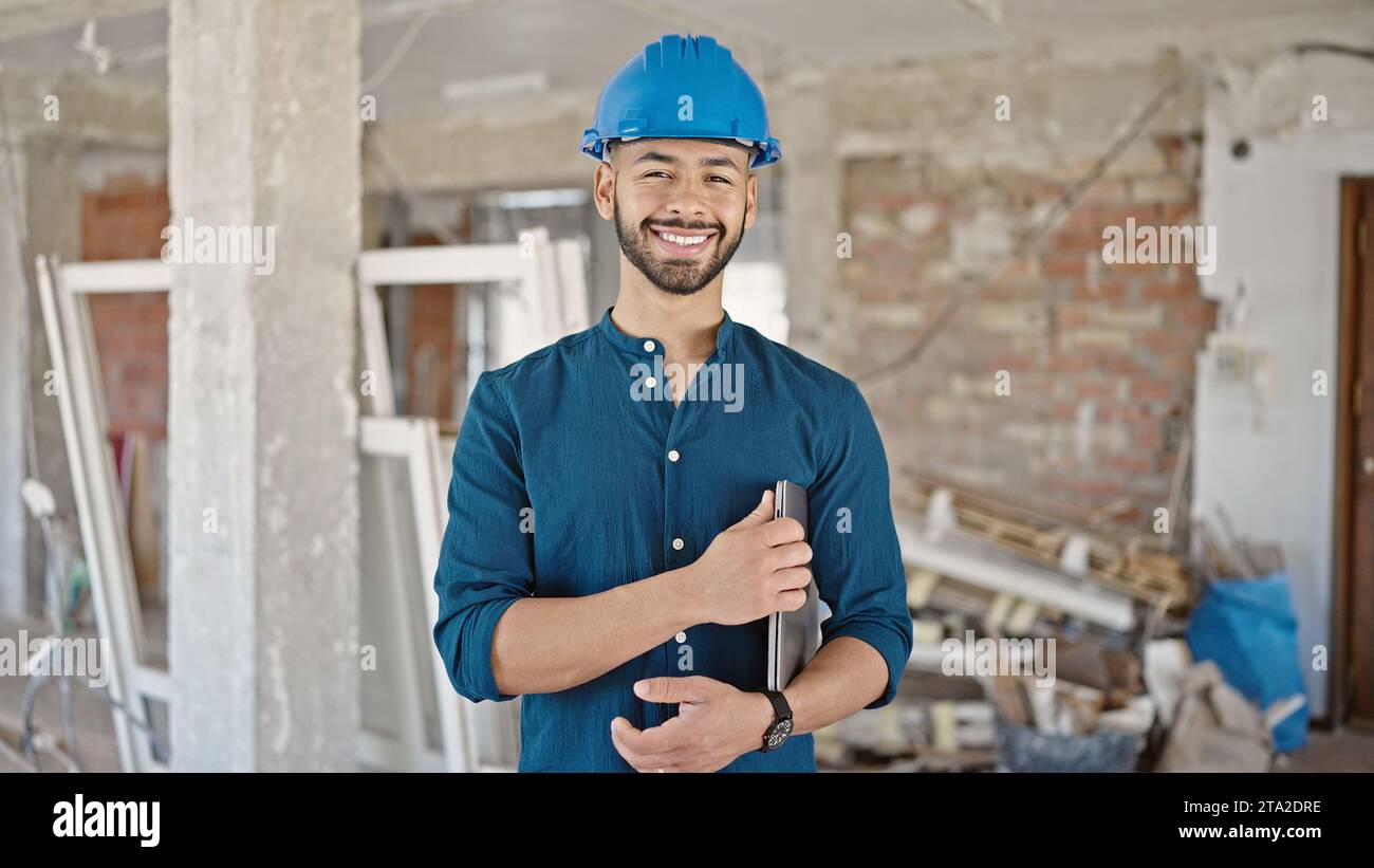 Young hispanic man architect wearing hardhat smiling holding laptop at construction site Stock ...