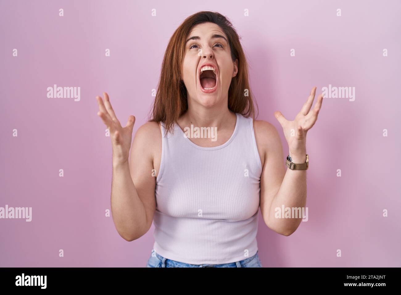 Brunette woman standing over pink background crazy and mad shouting and ...