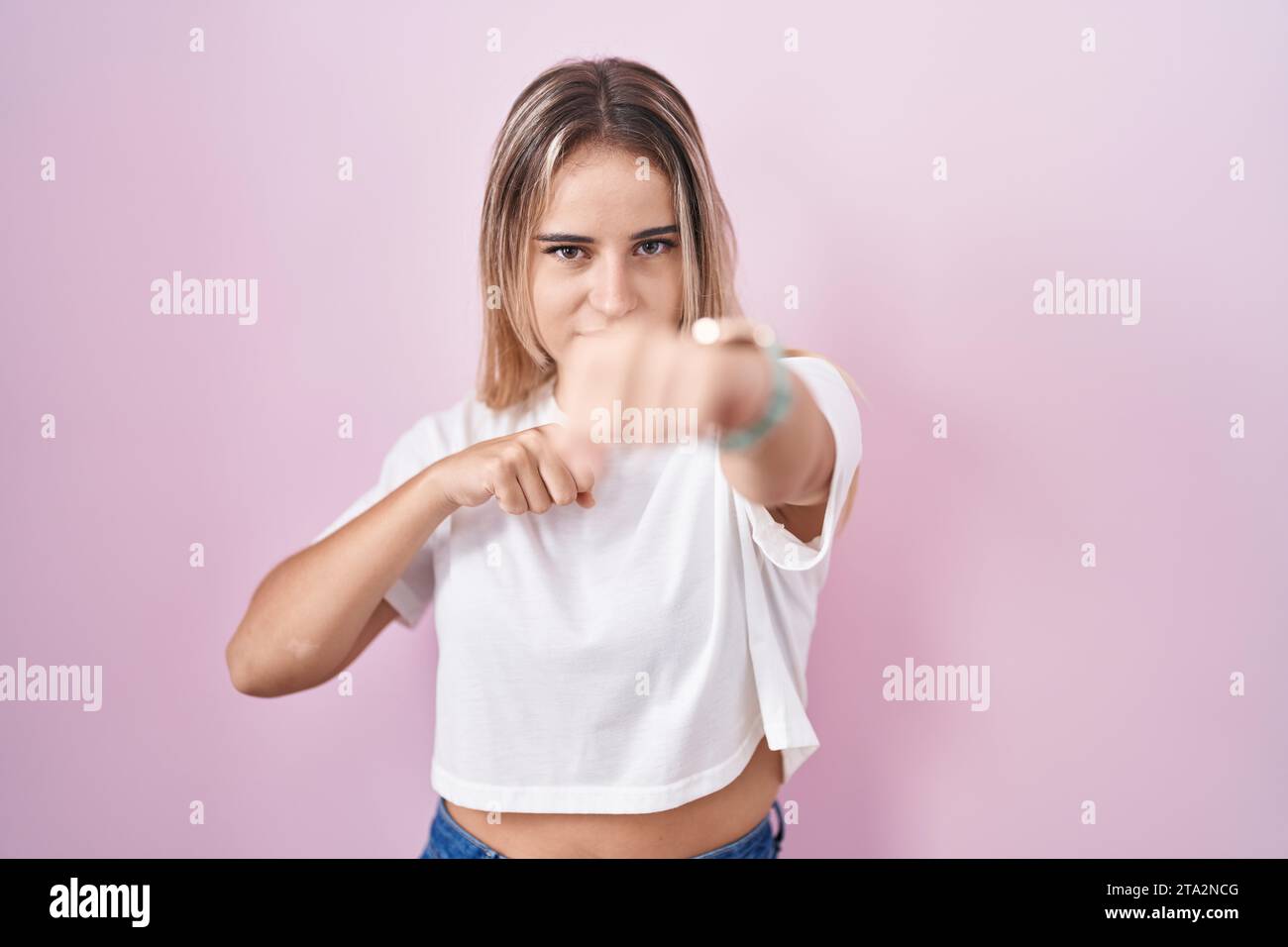 Young blonde woman standing over pink background punching fist to fight ...