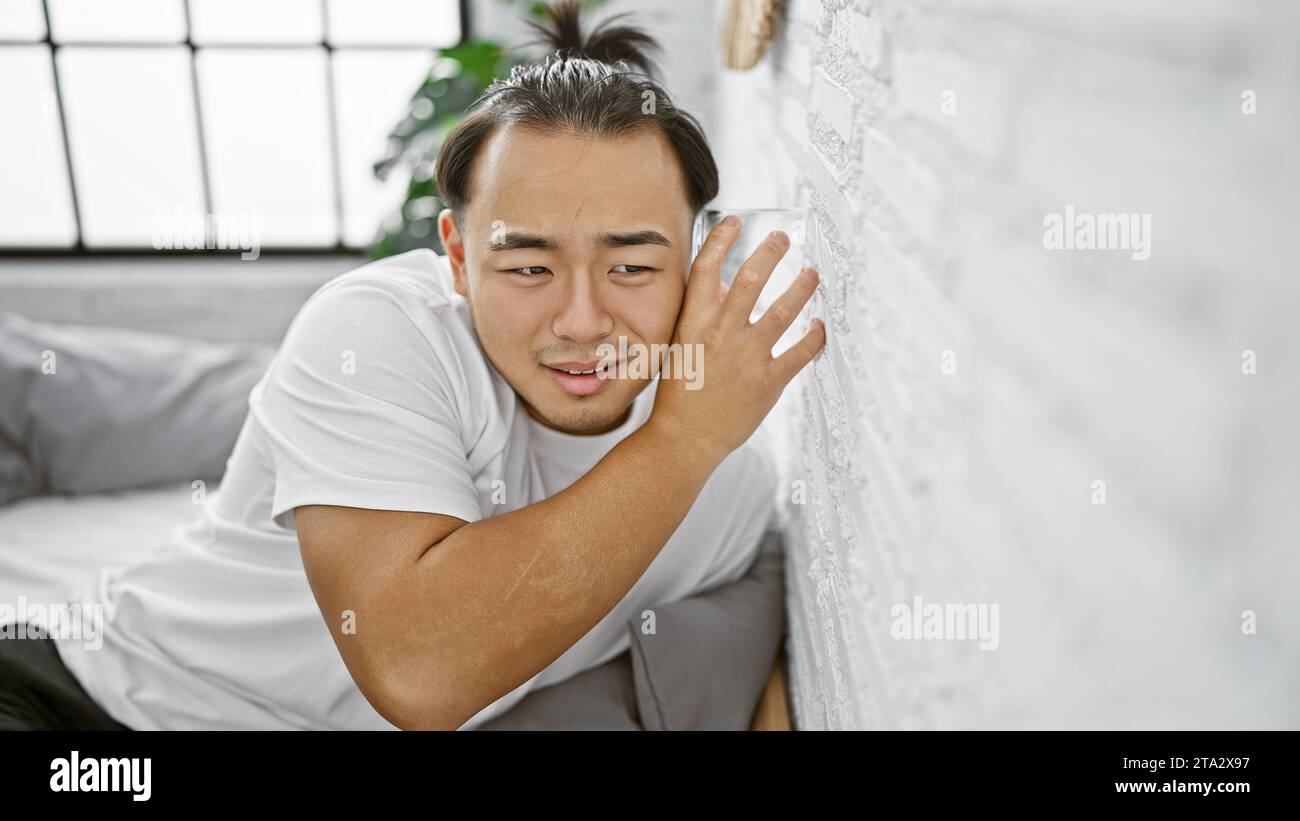 Nosy young chinese man sneakily listening through bedroom wall with glass, overcome by curiosity ...