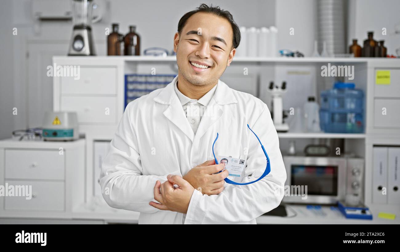 Confident young chinese scientist, smiling as he holds safety glasses ...
