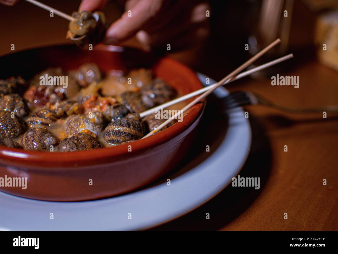 DETAIL VIEW OF SNAILS DISH IN SAUCE IN A CLAY POT. SOMEONE IS EATING ...