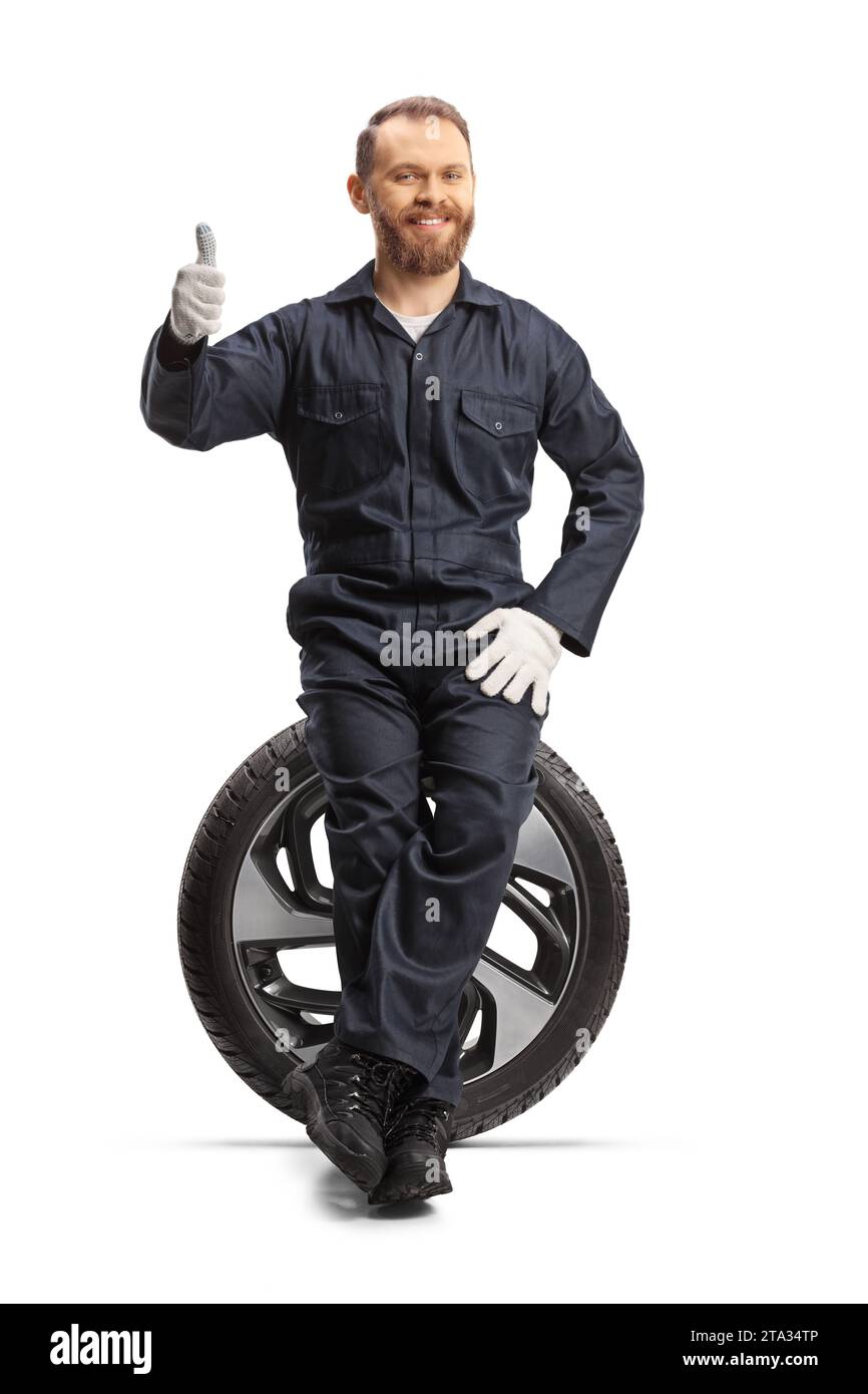 Car mechanic in a uniform gesturing thumbs up and sitting on a tire ...