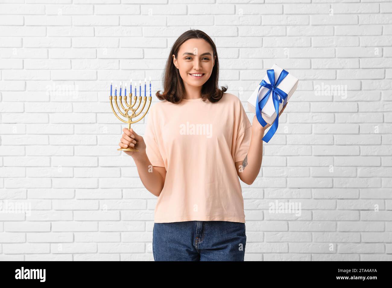 Pretty young woman with menorah and gift box on white brick wall background. Hanukkah ...