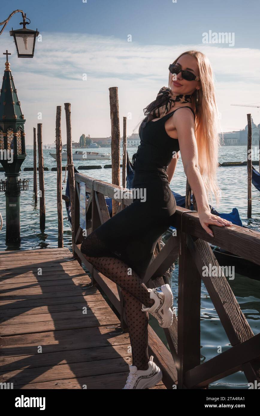 Blonde woman in black dress and white sneakers posing on wooden pier in ...
