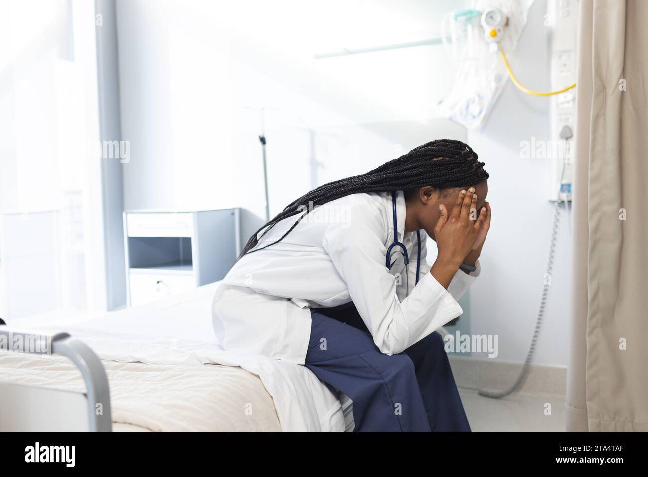 Tired african american female doctor wearing lab coat sitting on bed in ...