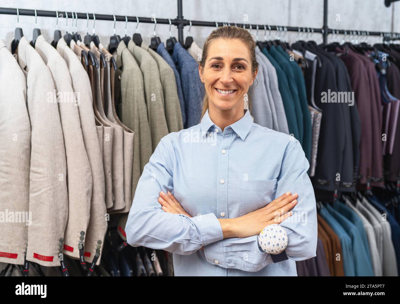 Smiling tailor with crossed arms in front of a rack of suits in a ...