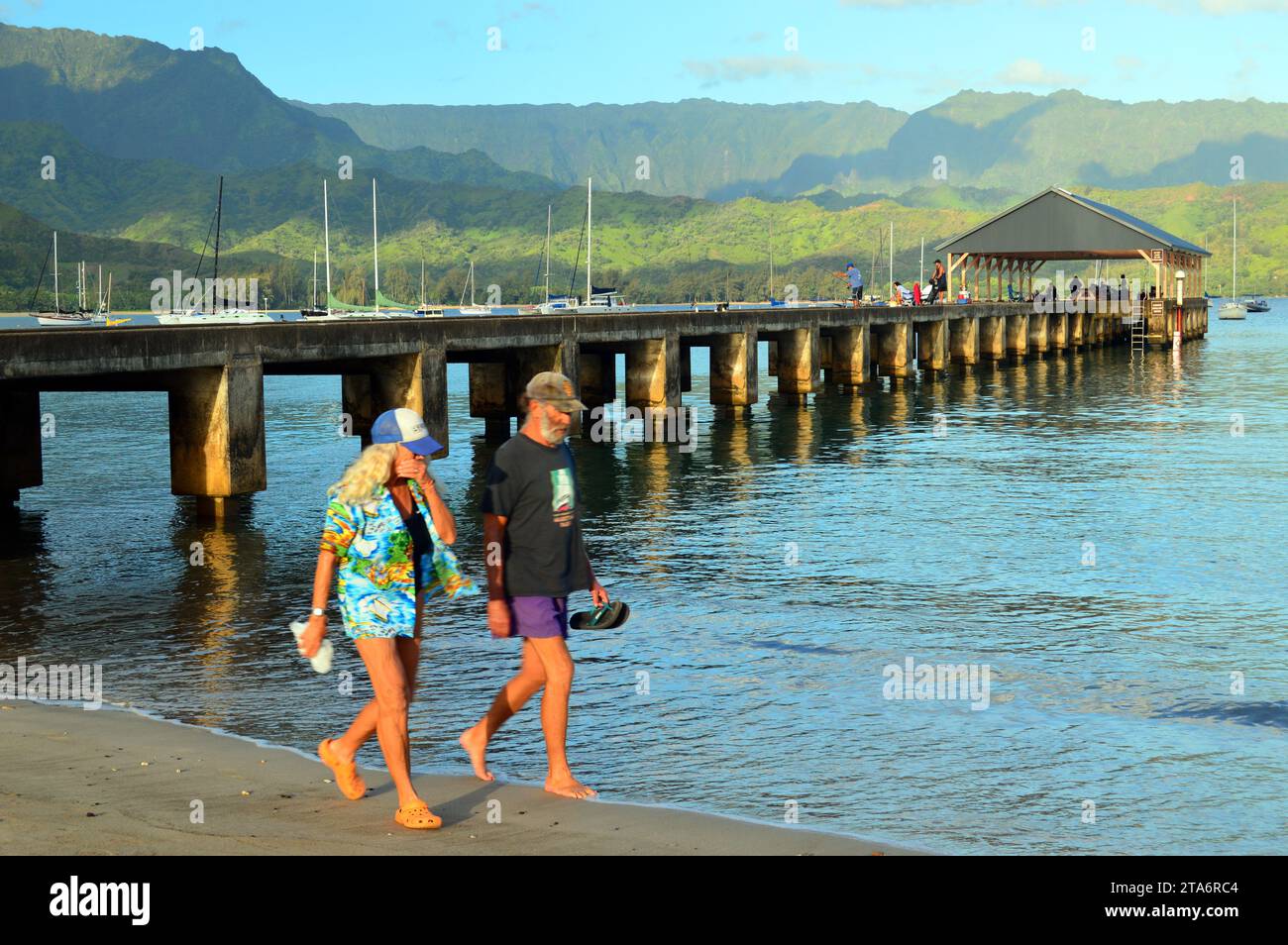An adult couple walks on the beach during their vacation, enjoying the Hanalei Pier on Kauai ...