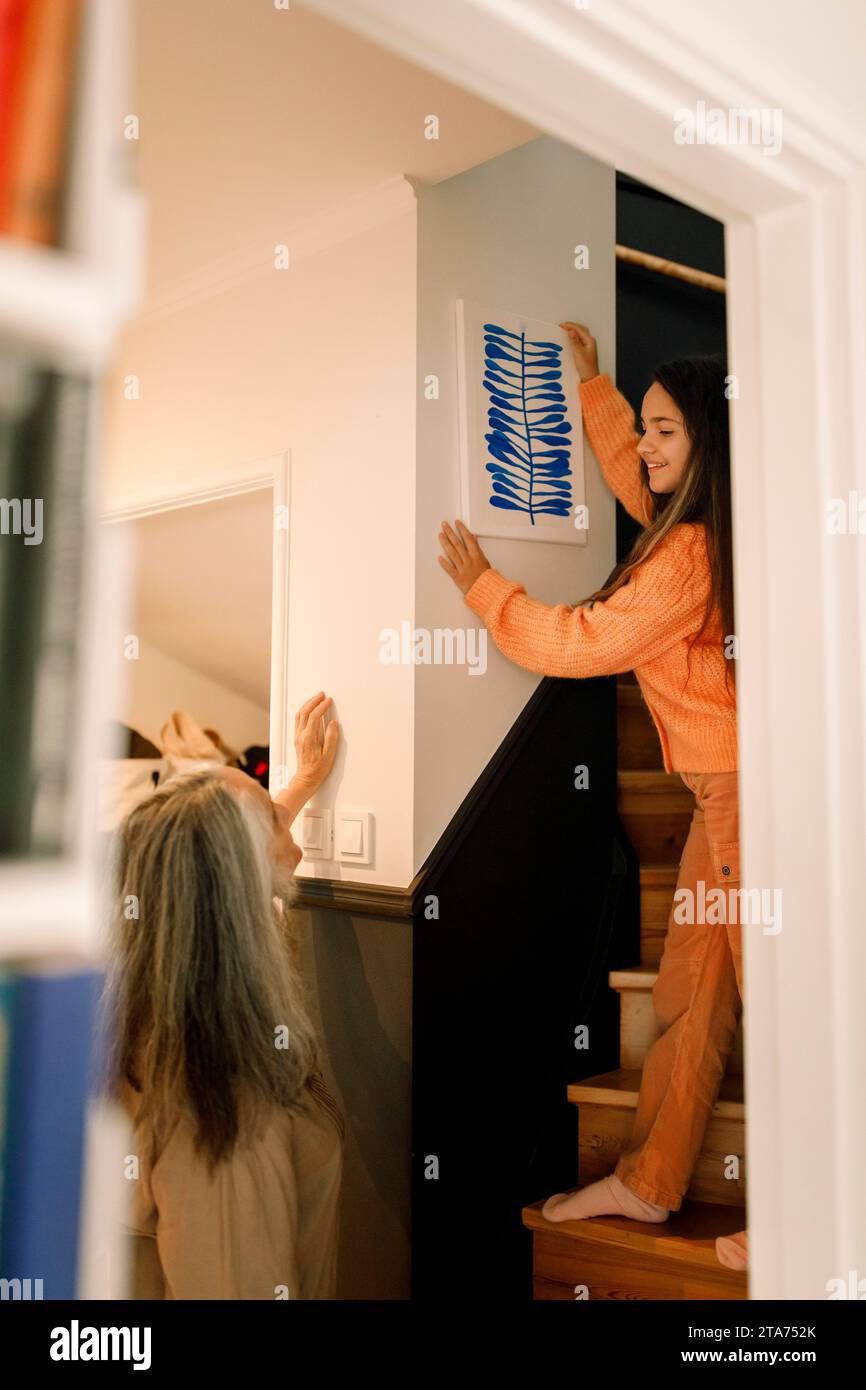 Girl arranging painting on wall standing on steps by grandmother at ...