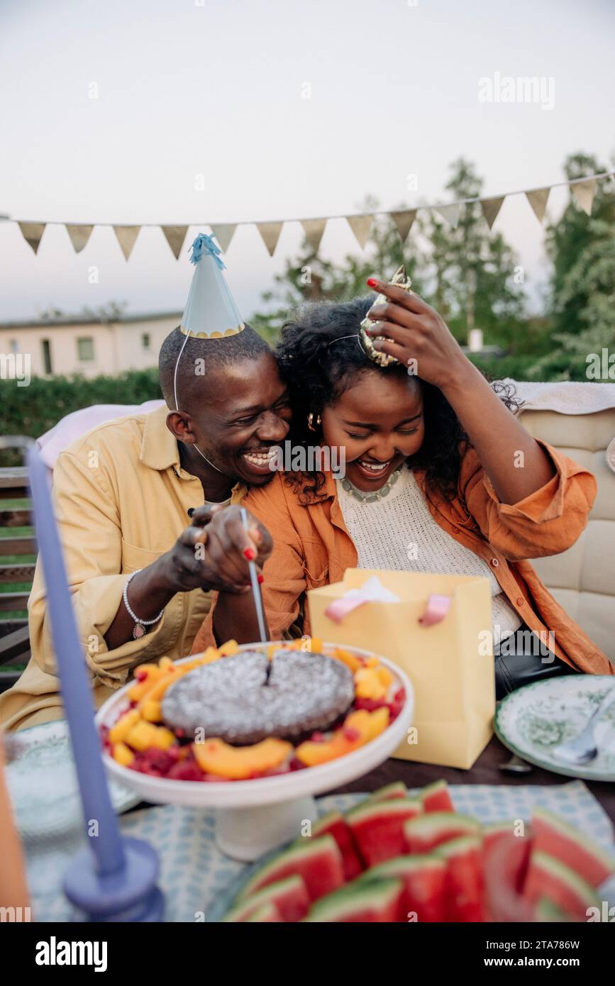 Happy young man sitting with female friend cutting cake while ...