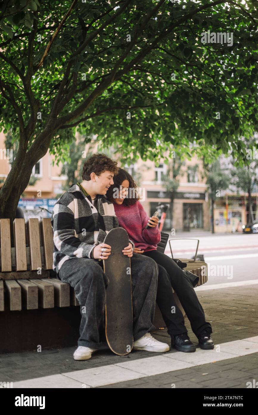 Smiling girl sharing smart phone with male teenage friend sitting on ...