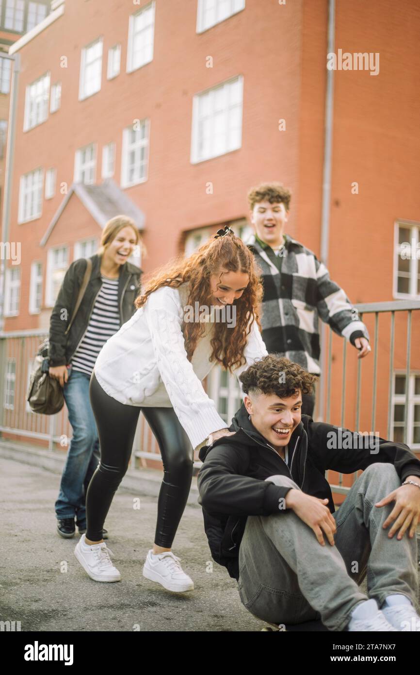 Teenage girl pushing male friend sitting on skateboard Stock Photo - Alamy