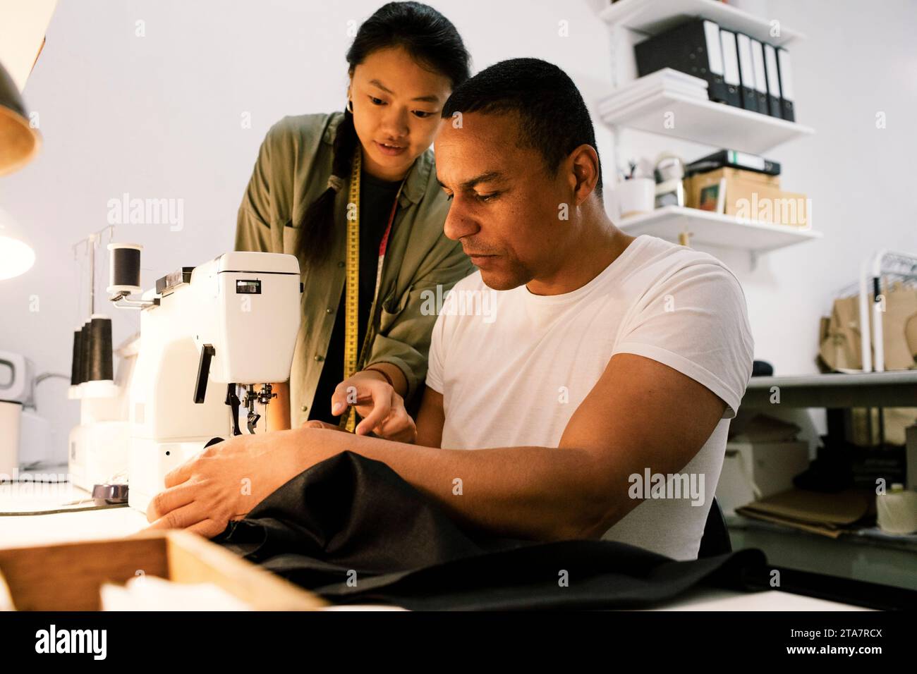 Female colleague assisting male tailor stitching clothes using sewing ...