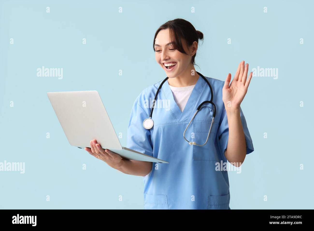 Beautiful female doctor with laptop waving hand on blue background ...