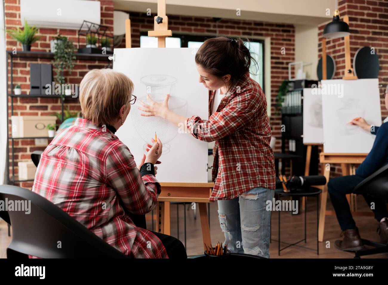 Female art teacher standing near easel explaining senior woman how to ...