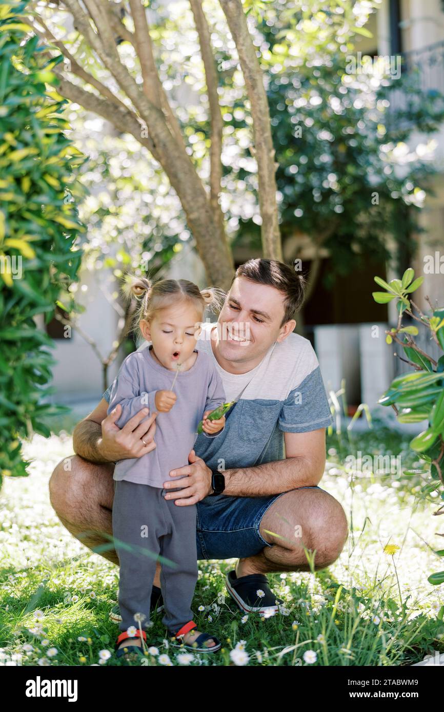 Smiling dad squatting near little girl blowing on dandelion in garden Stock Photo - Alamy