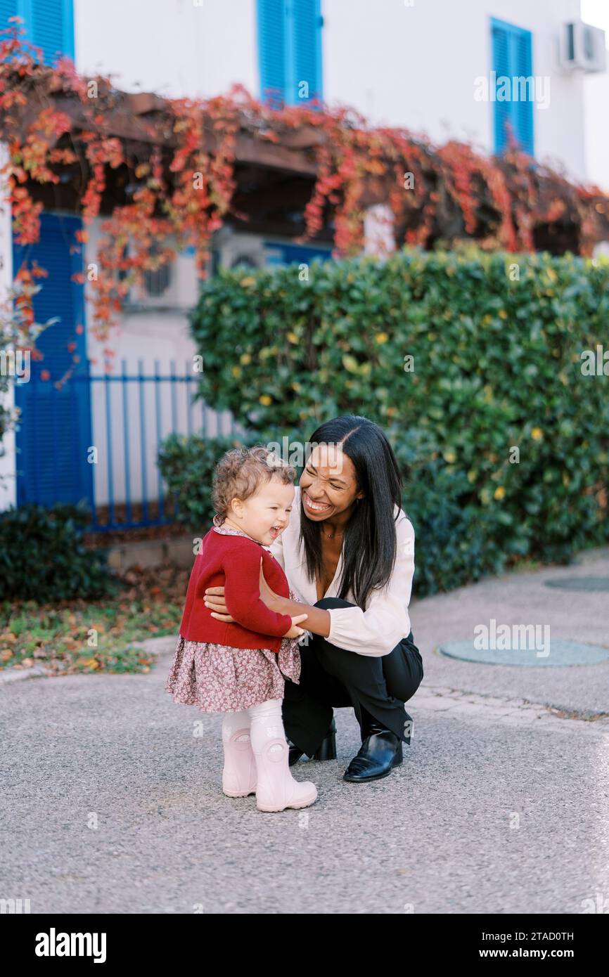 Laughing mother hugging the waist of a smiling little girl crouching ...